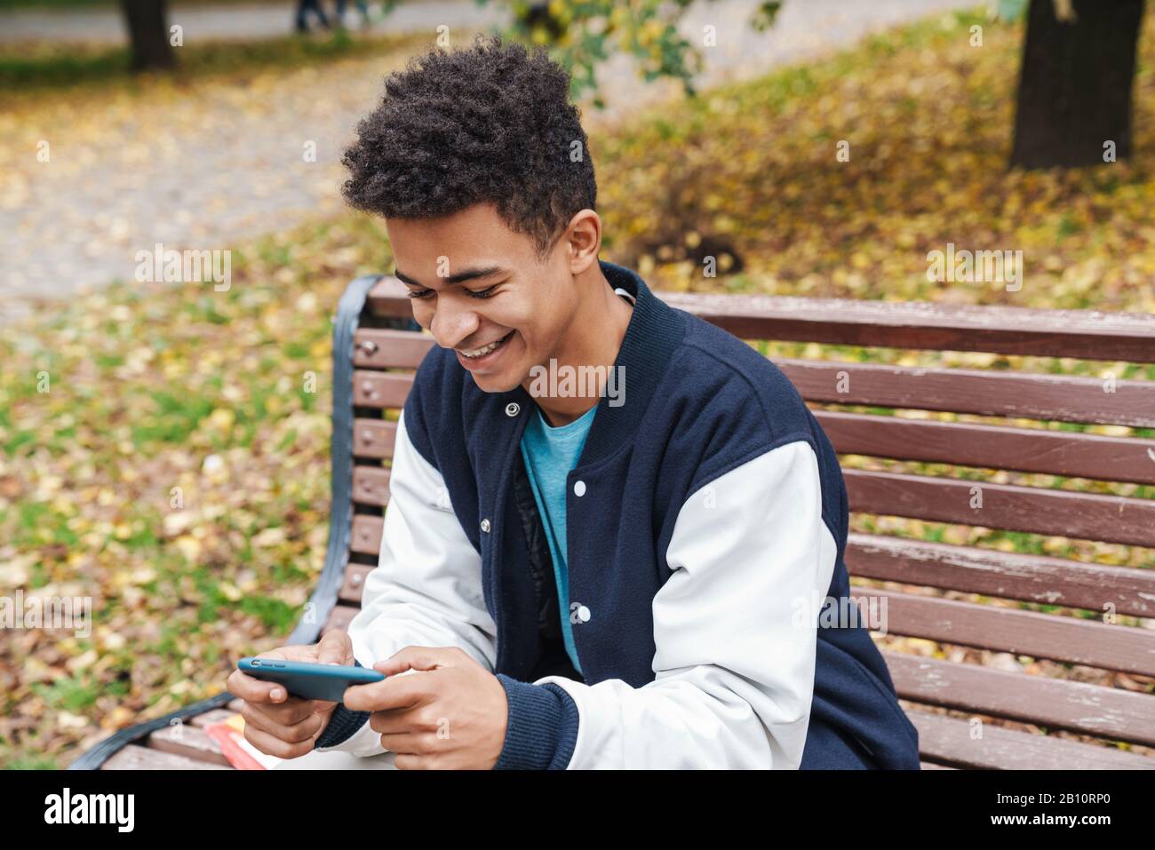 Smiling african boy student sitting on a bench at the park, using ...