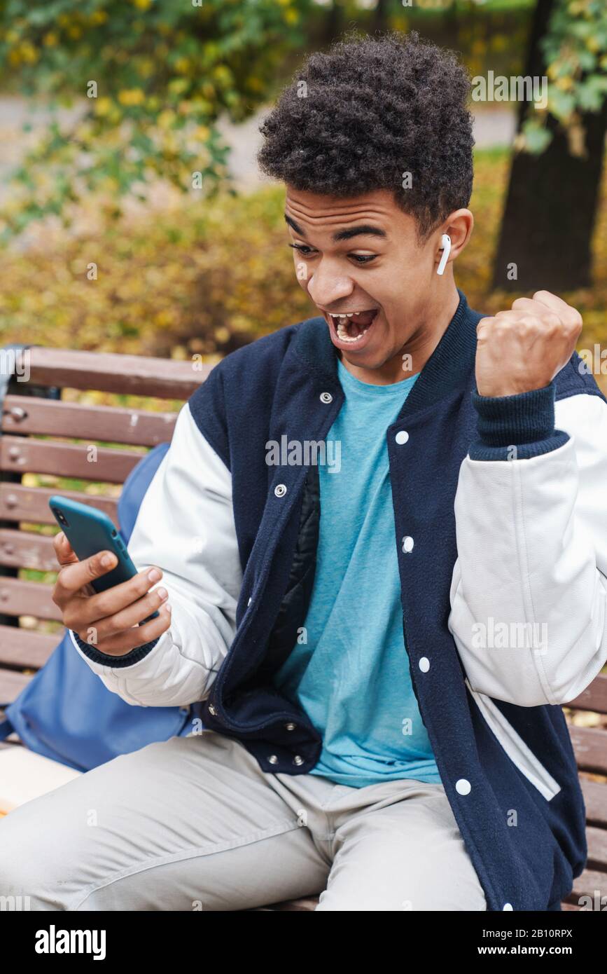 Excited african boy student sitting on a bench at the park, playing ...
