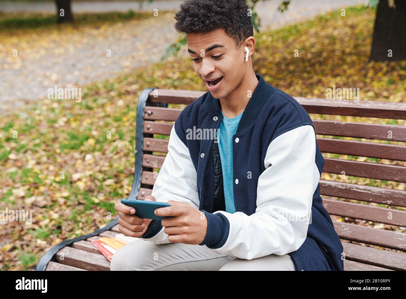 Excited african boy student sitting on a bench at the park, playing ...