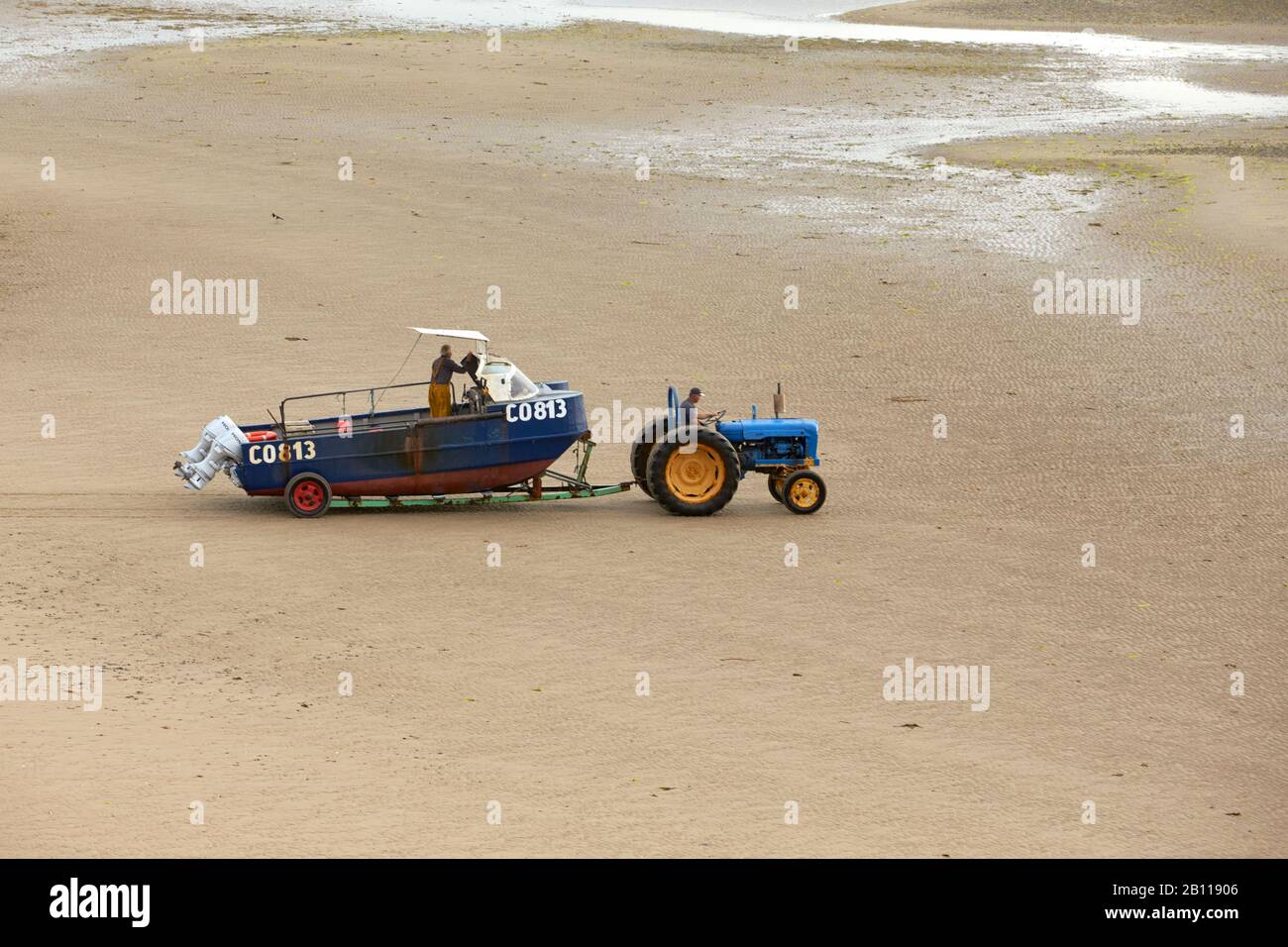 Blue tractor with yellow wheels towing trailer with fishing boat across ...