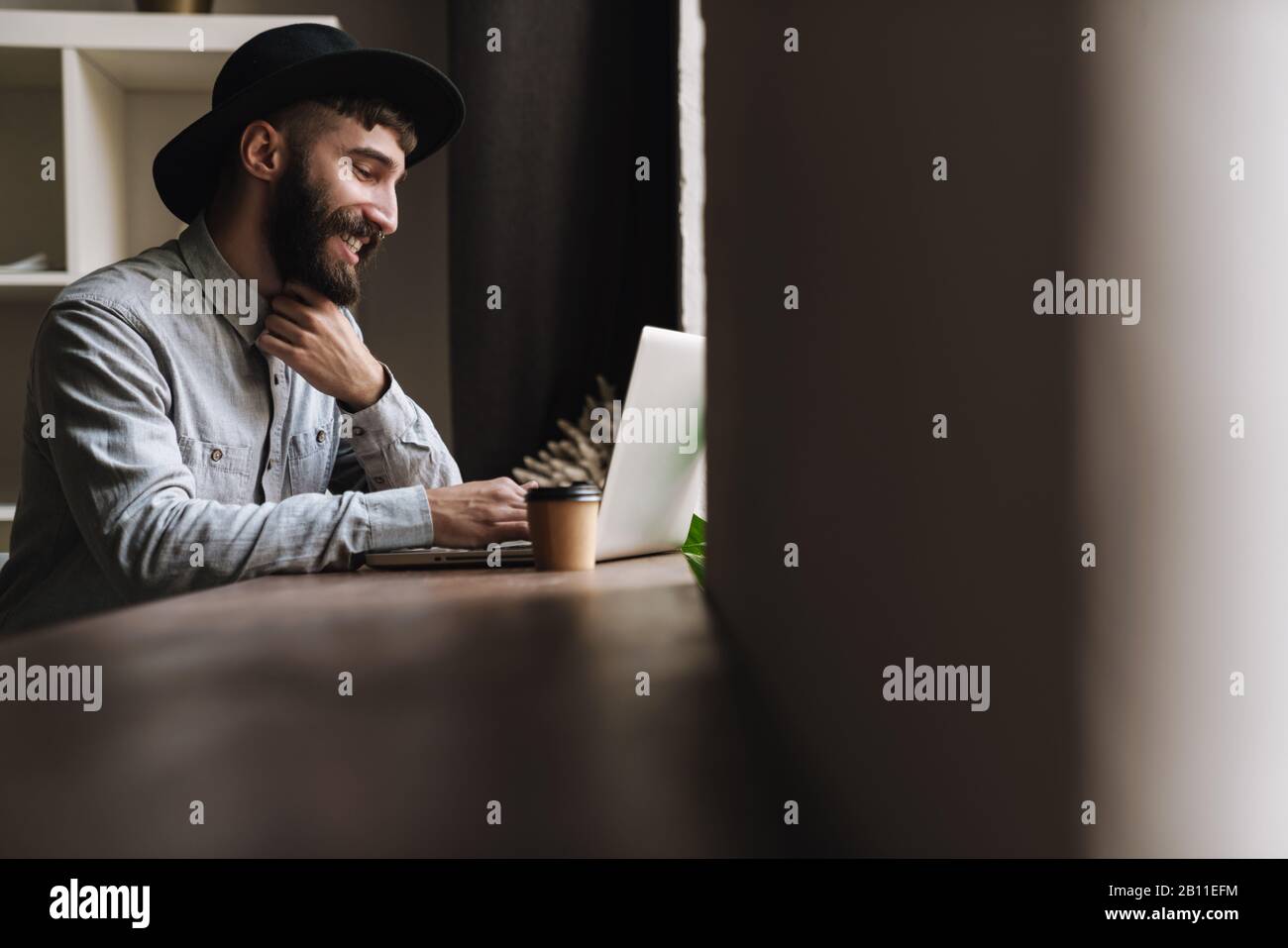 Photo of laughing young man wearing hat typing on laptop and drinking ...