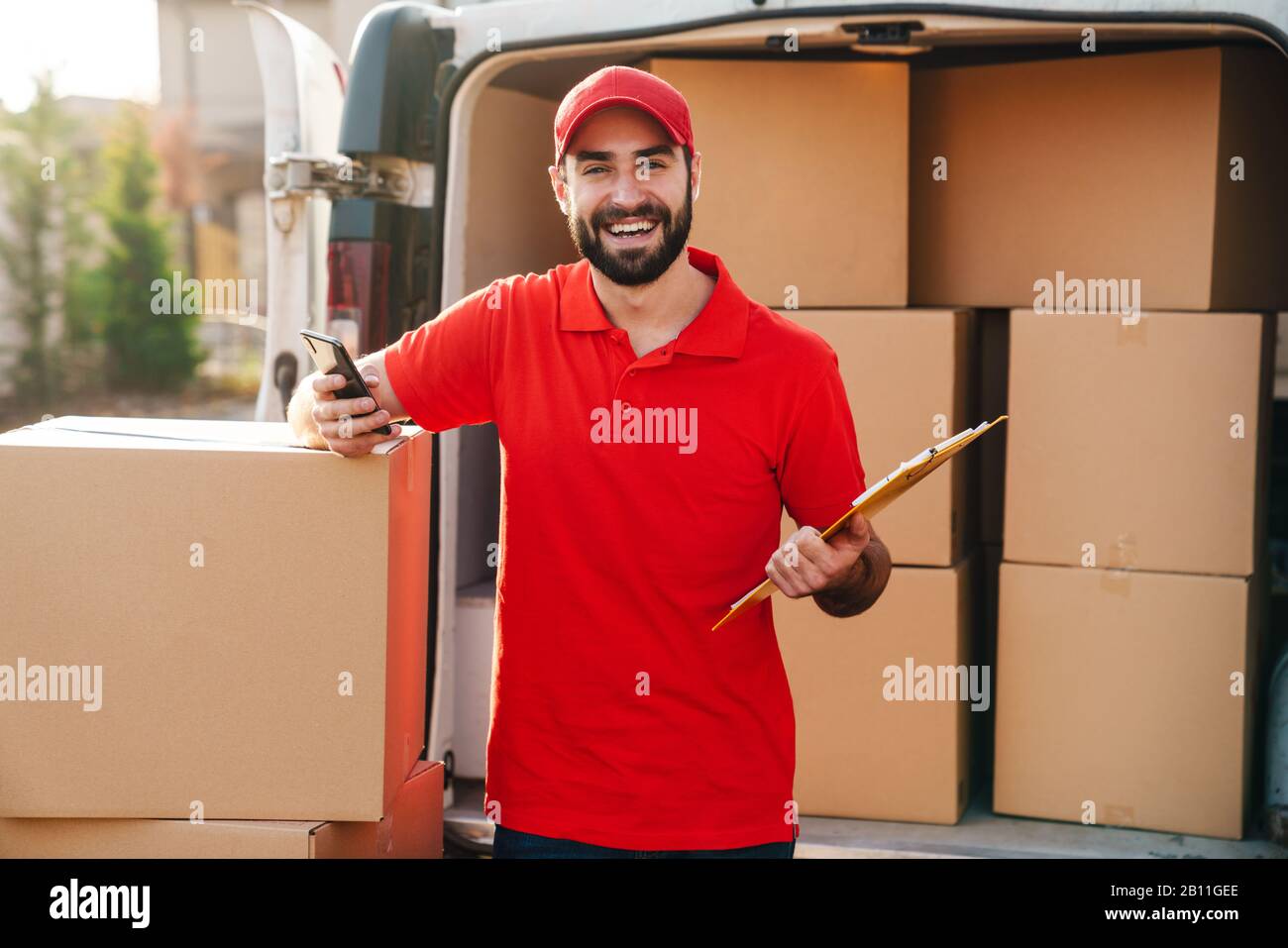 Image of happy delivery man holding clipboard and using cellphone while ...