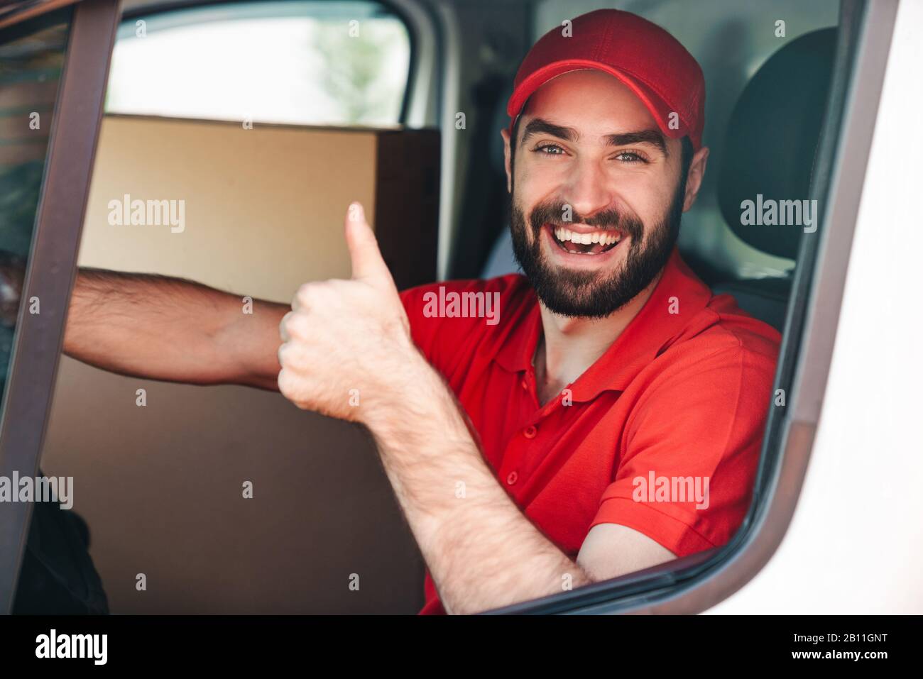 Image of happy young delivery man in red uniform showing thumb up and ...