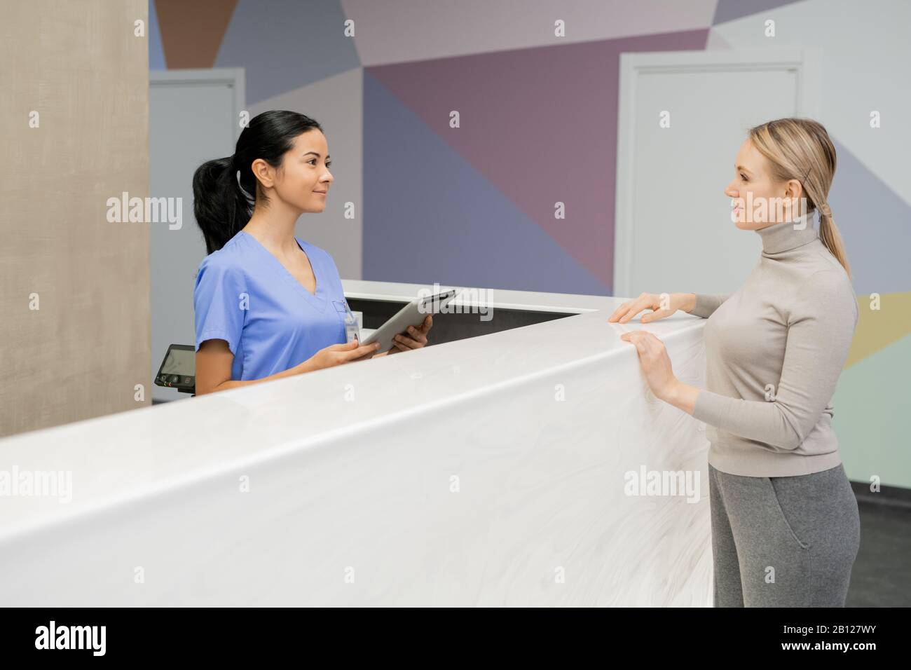 Young female assistant standing by reception counter and consulting ...