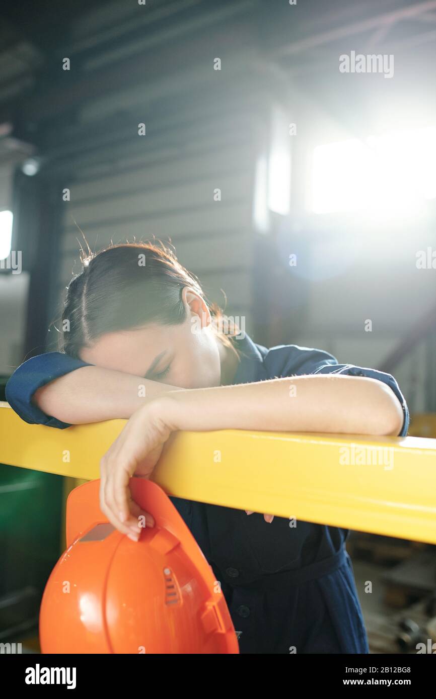 Exhausted young engineer in blue coveralls holding hardhat while taking ...