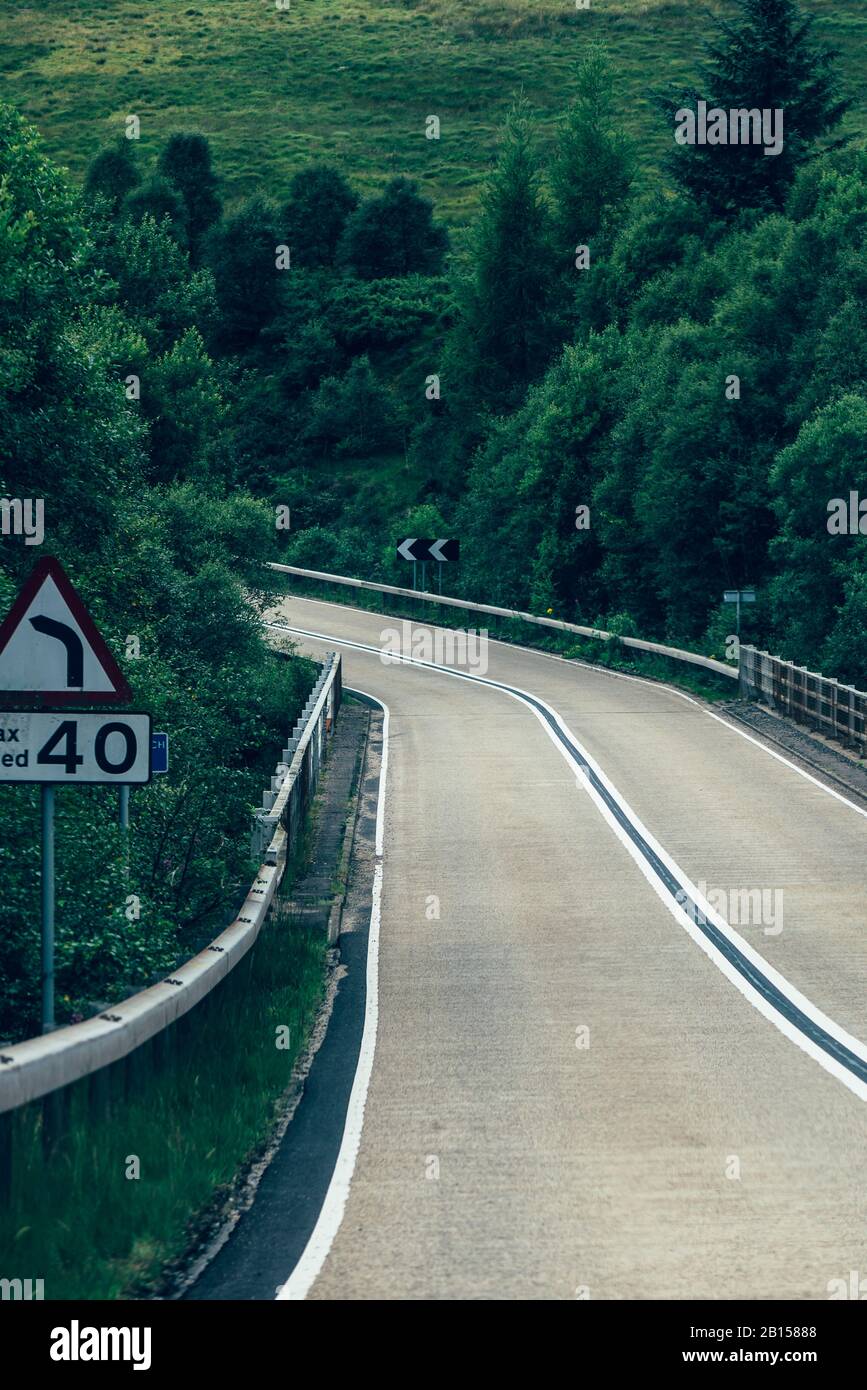 Winding rural road leading through green hills and forest in Scotland ...