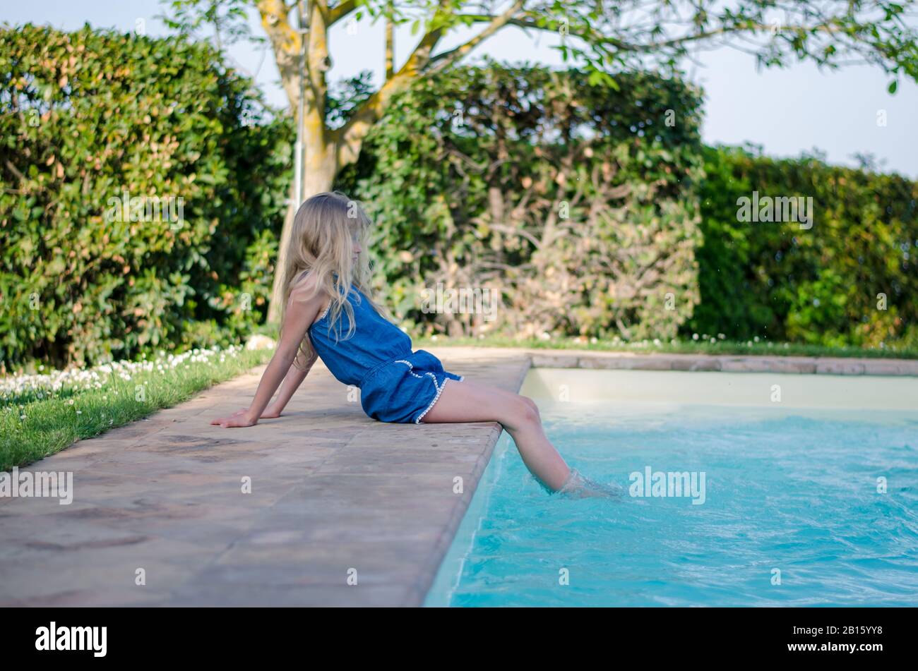 adorable little child sitting by the swimming pool among green trees ...