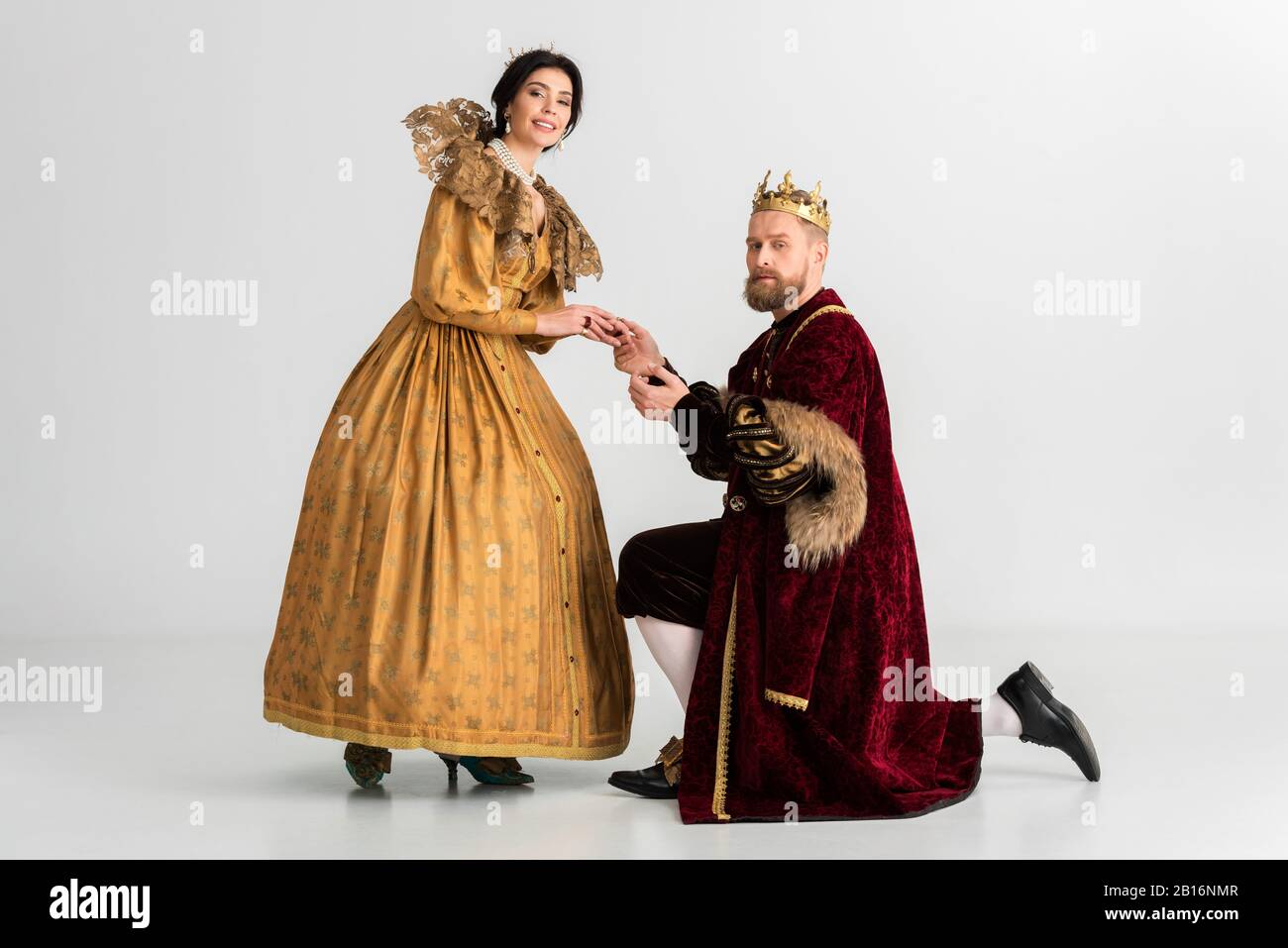 king with crown bending on knee and holding hand of smiling queen on ...