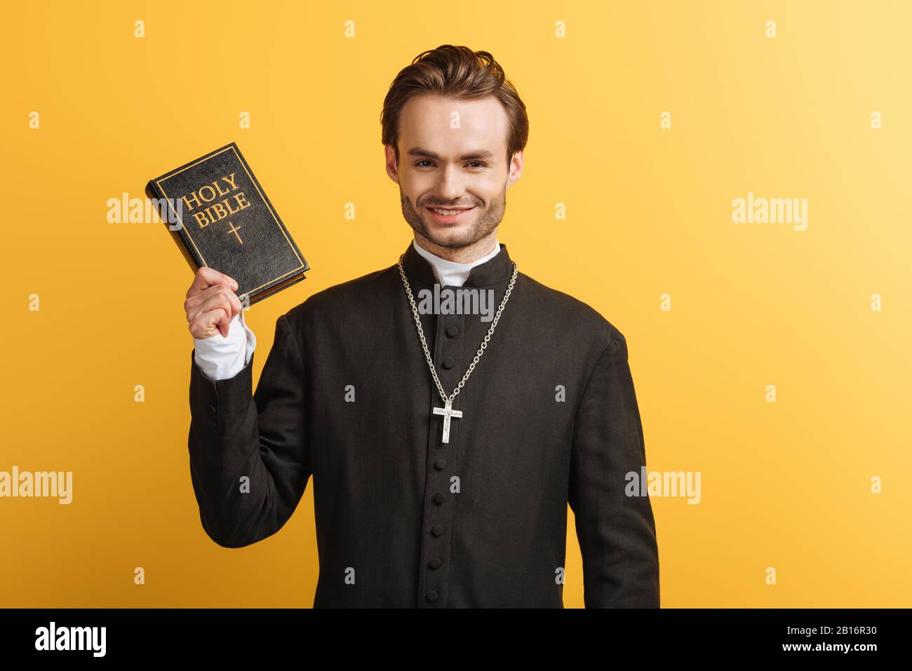 happy catholic priest holding bible while smiling at camera isolated on ...