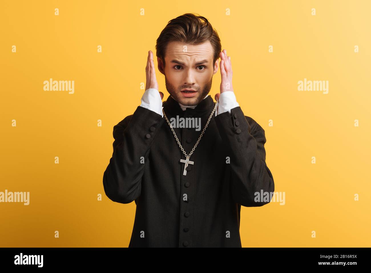 shocked catholic priest holding hands near head and looking at camera ...