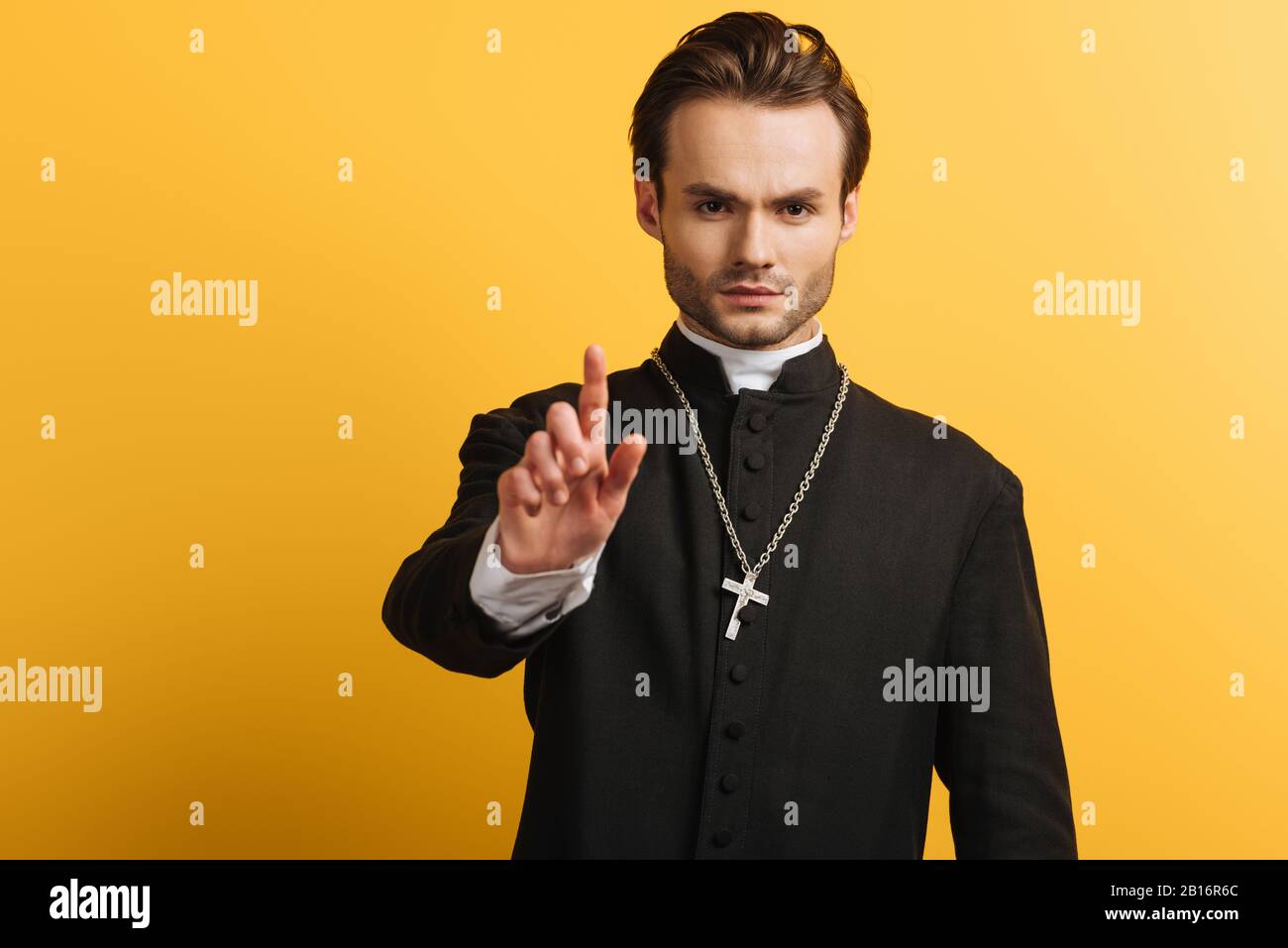 serious catholic priest showing warning gesture isolated on yellow ...