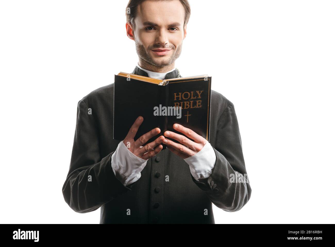 young, smiling catholic priest holding holy bible while looking at ...
