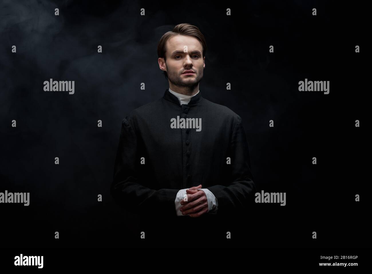 young serious catholic priest standing with folded hands on black ...