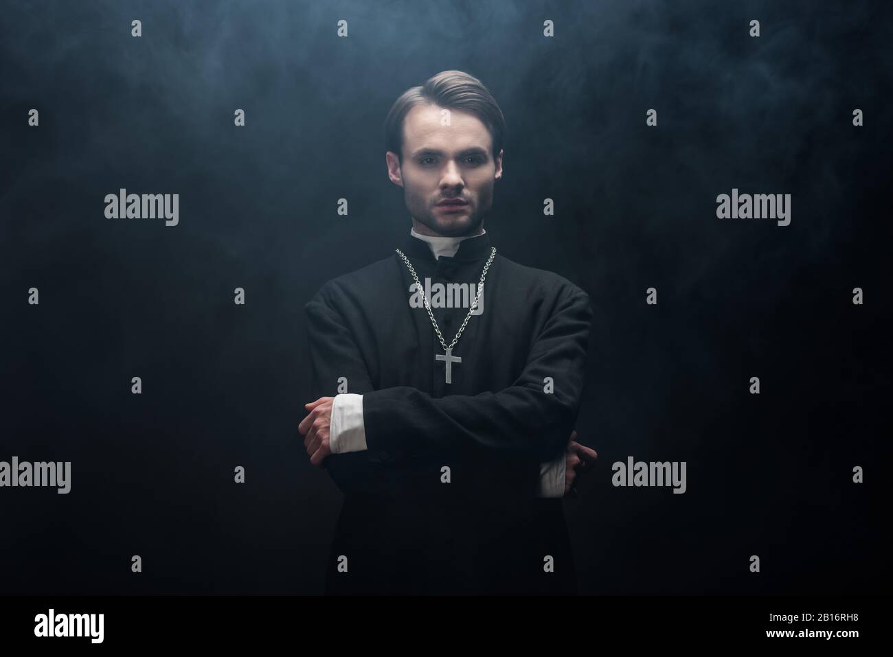 young serious catholic priest standing with crossed arms on black ...