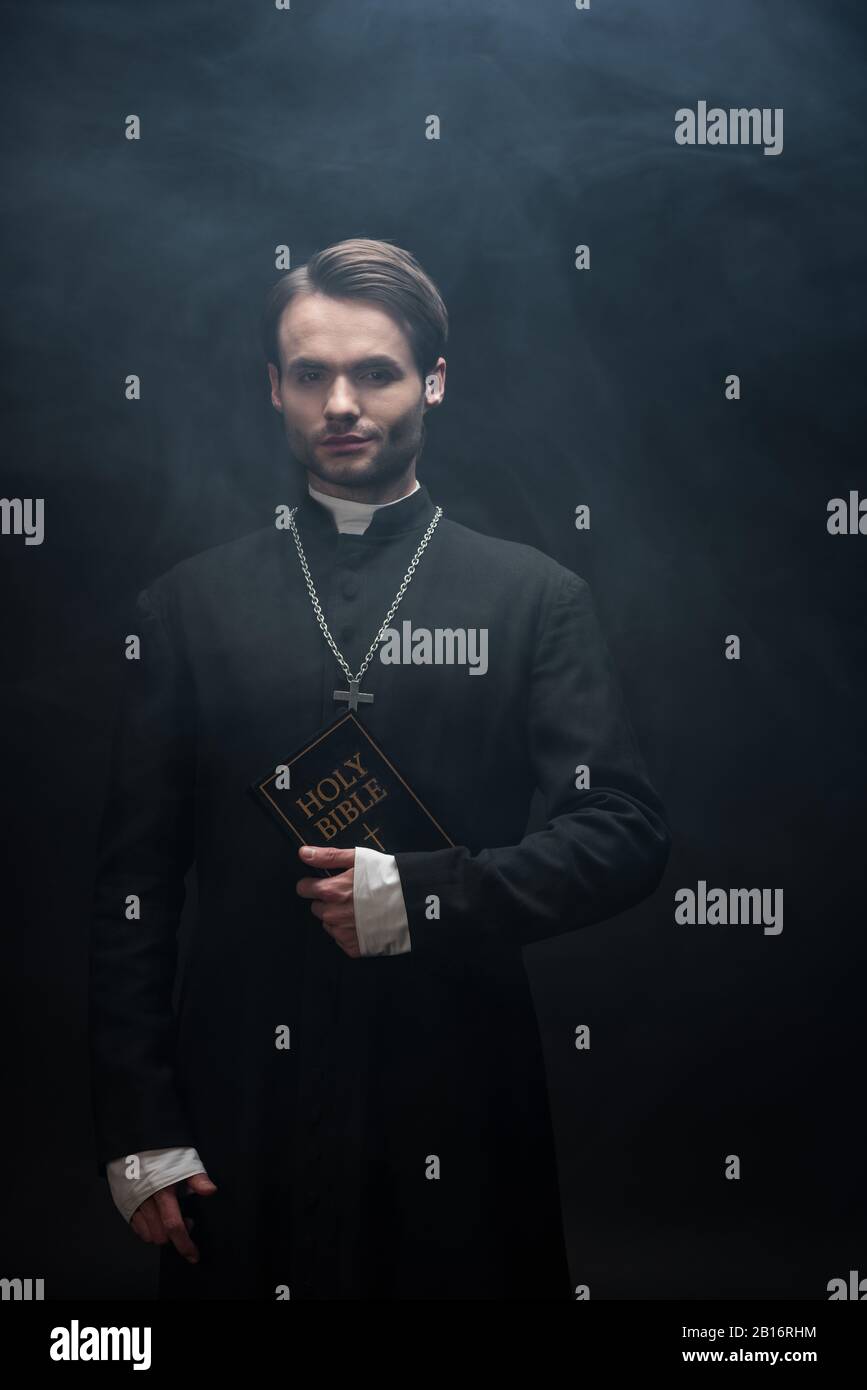 confident catholic priest holding holy bible and looking at camera on ...
