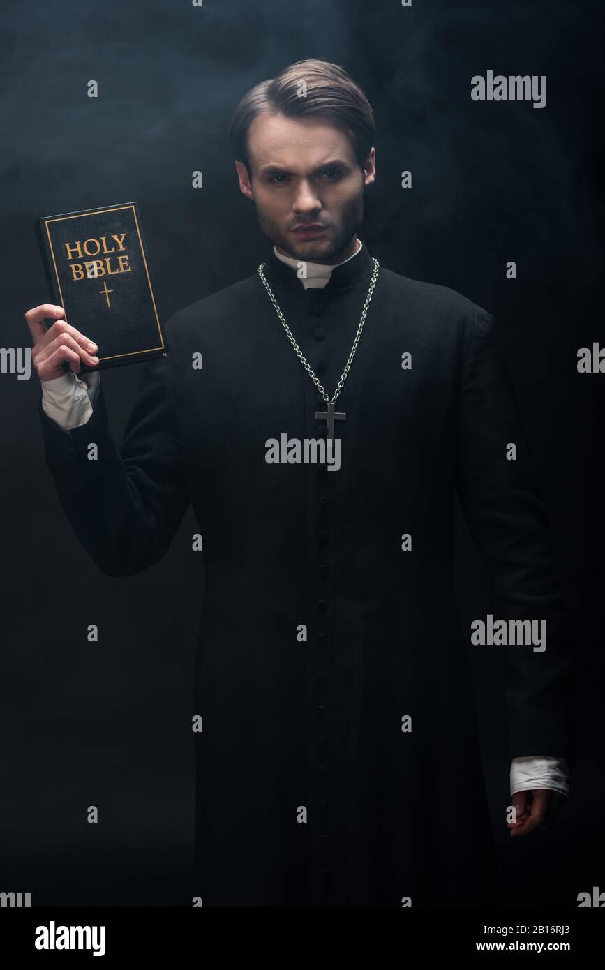young strict catholic priest showing holy bible at camera on black ...
