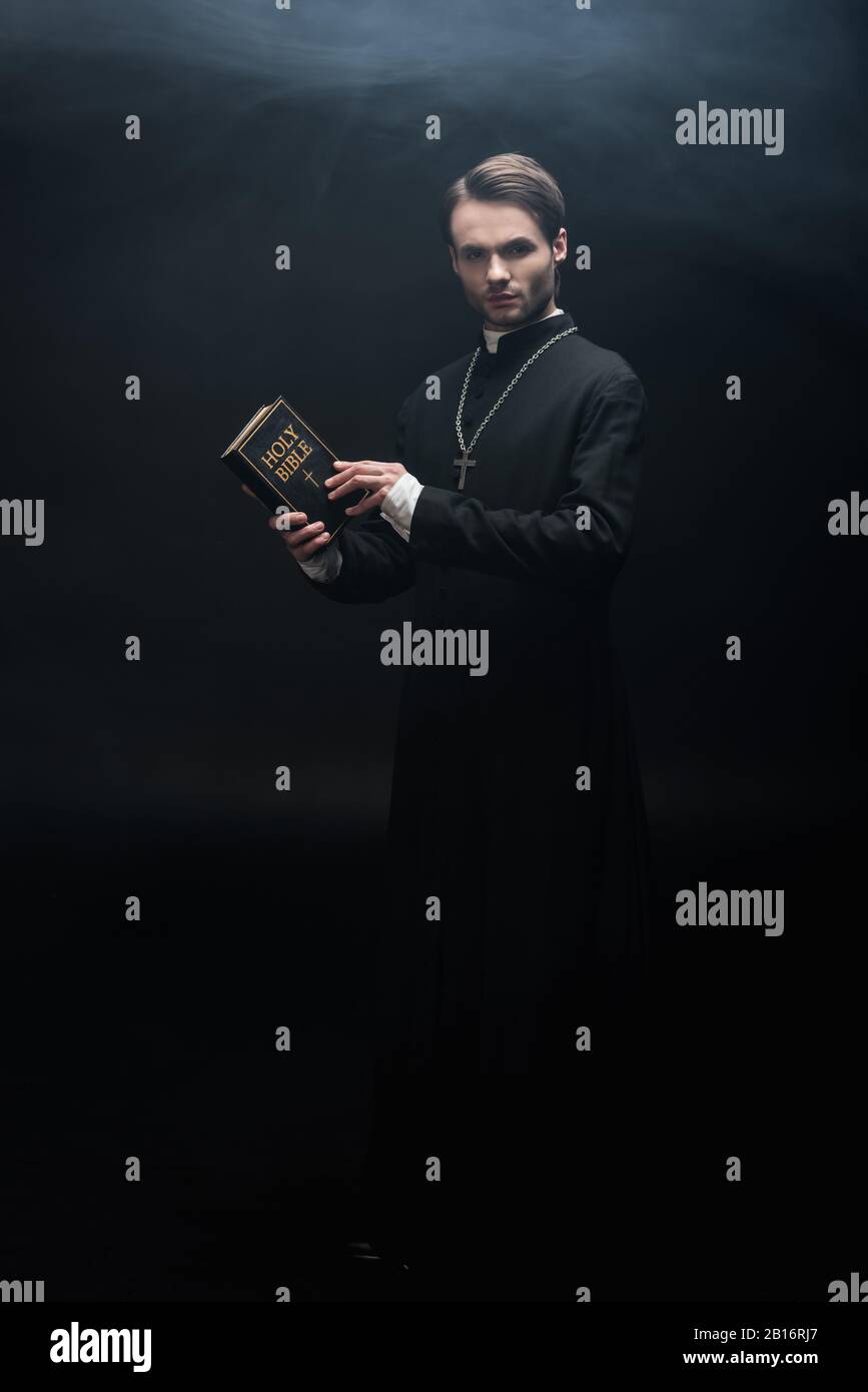 full length view of strict catholic priest holding holy bible and ...