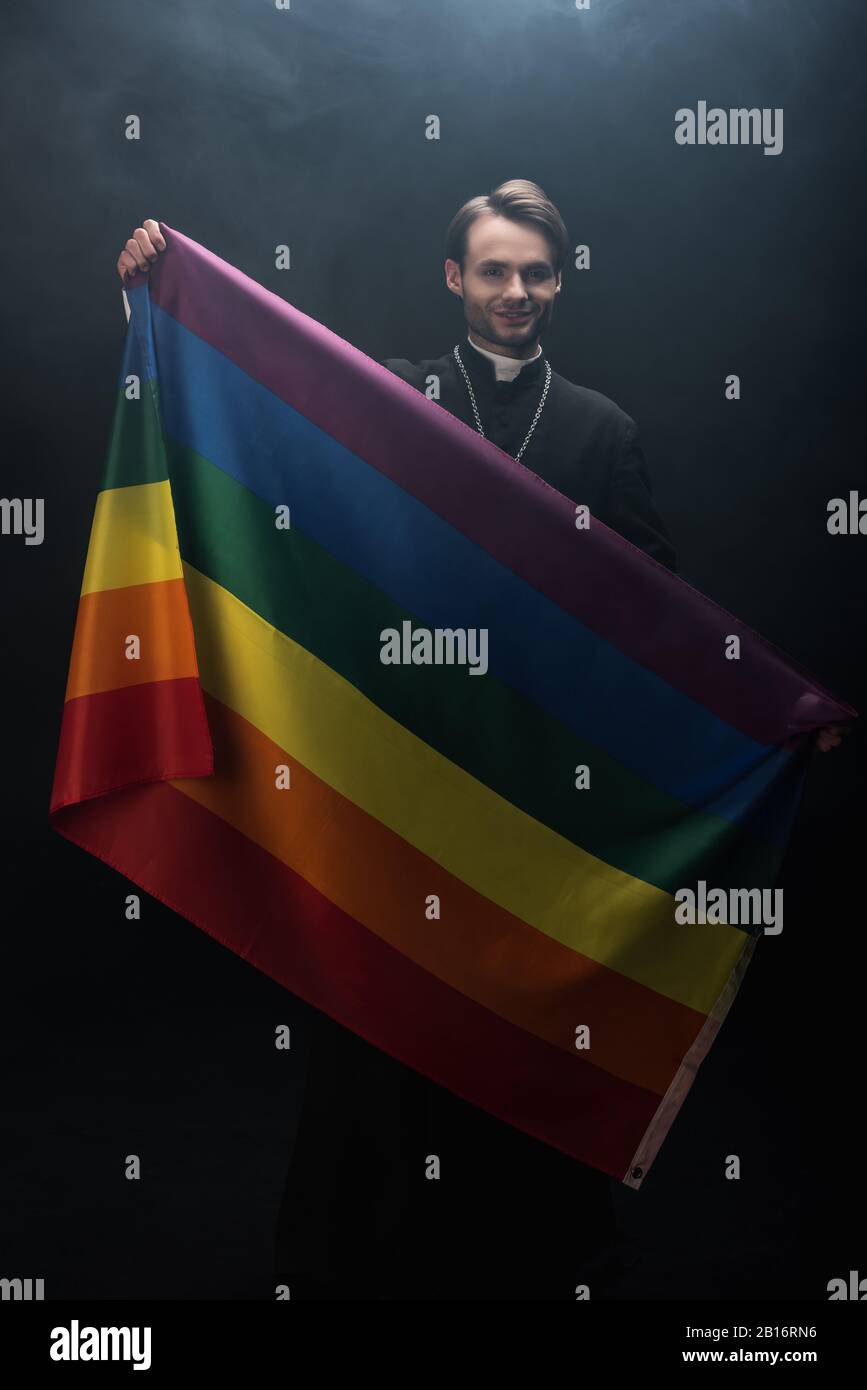 smiling catholic priest holding lgbt flag while looking at camera on ...