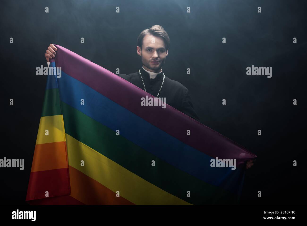smiling catholic priest holding lgbt flag while looking at camera on ...
