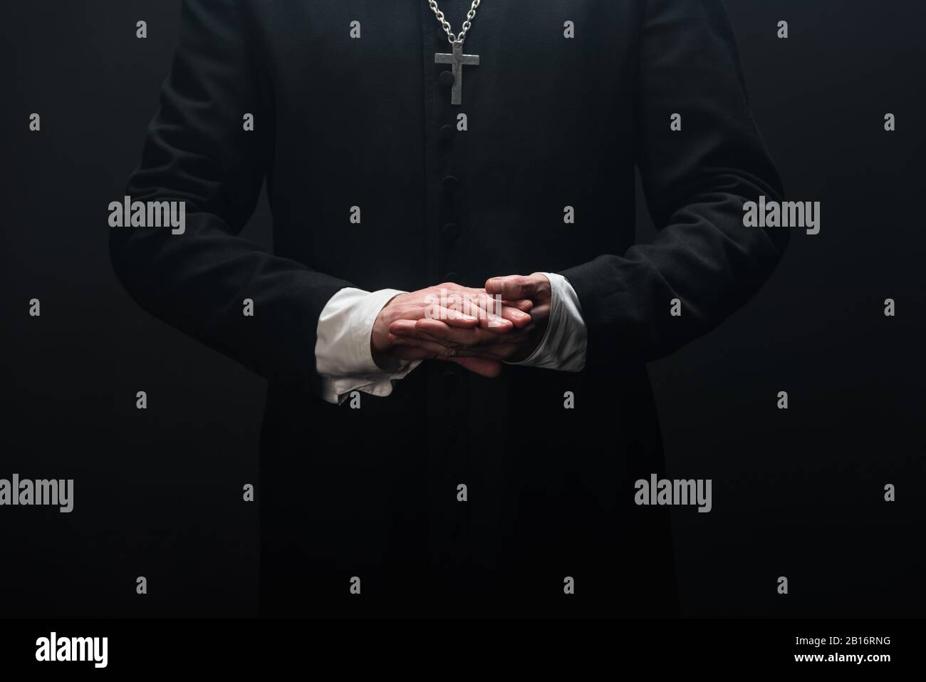 cropped of catholic priest standing with folded hands isolated on black ...