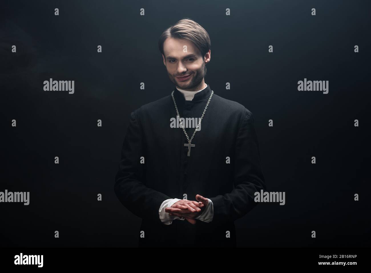 young smiling catholic priest standing with folded hands and looking at ...