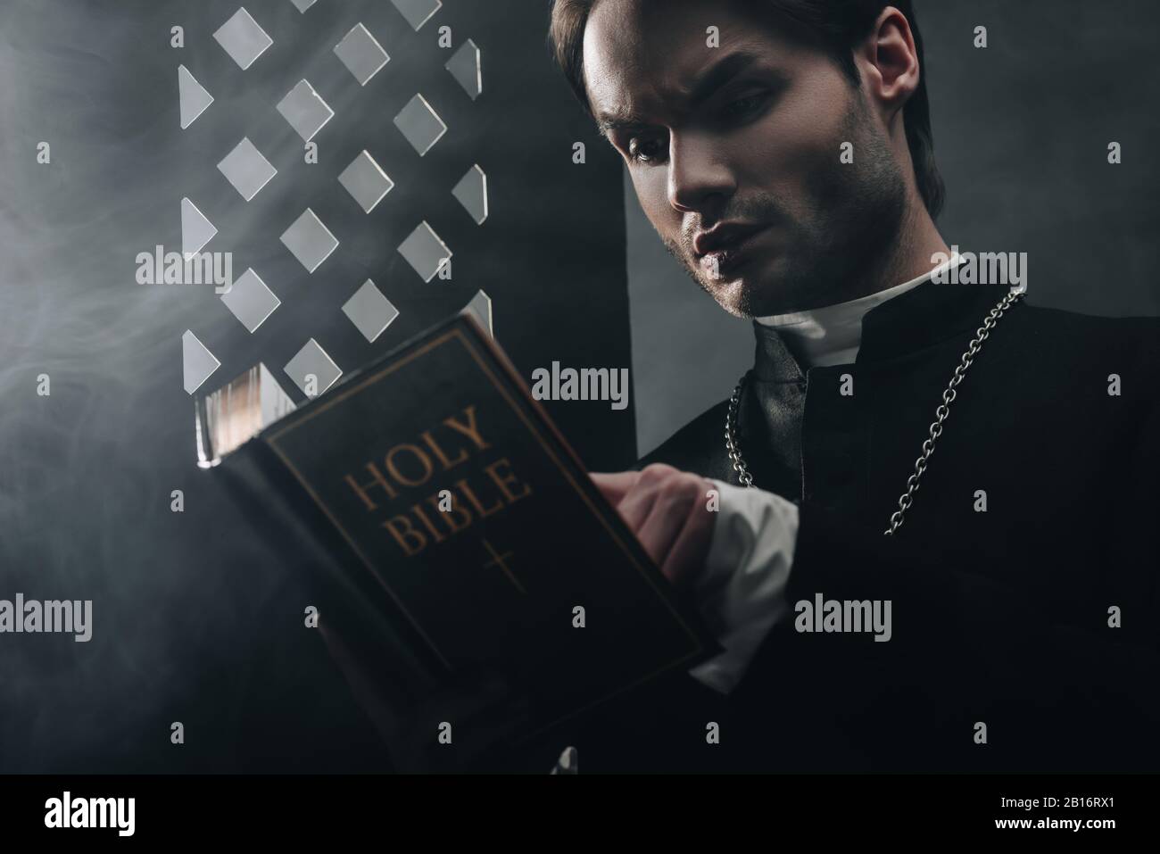 young thoughtful catholic priest reading bible near confessional grille ...