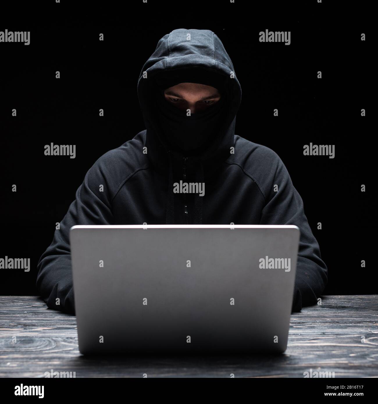 young hacker in mask and hood using laptop isolated on black Stock ...