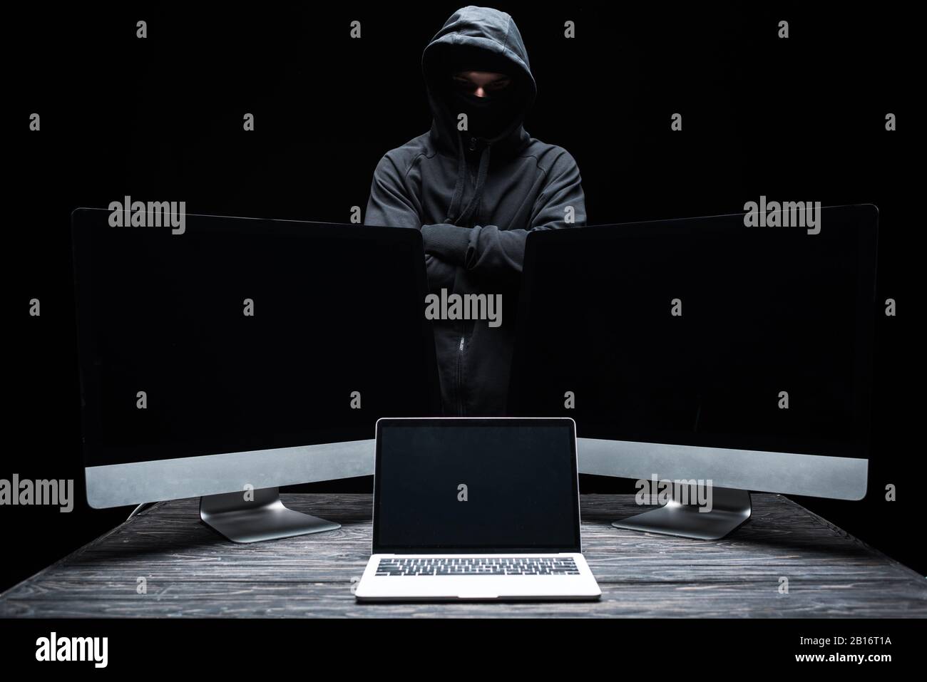 hacker in mask standing with crossed arms near computer monitors and ...
