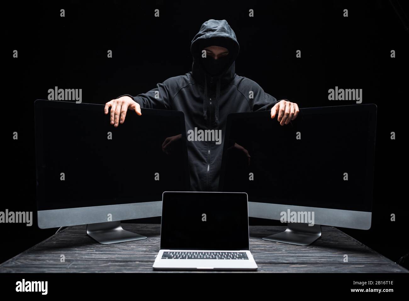 hacker in mask touching computer monitors near laptop with blank screen ...