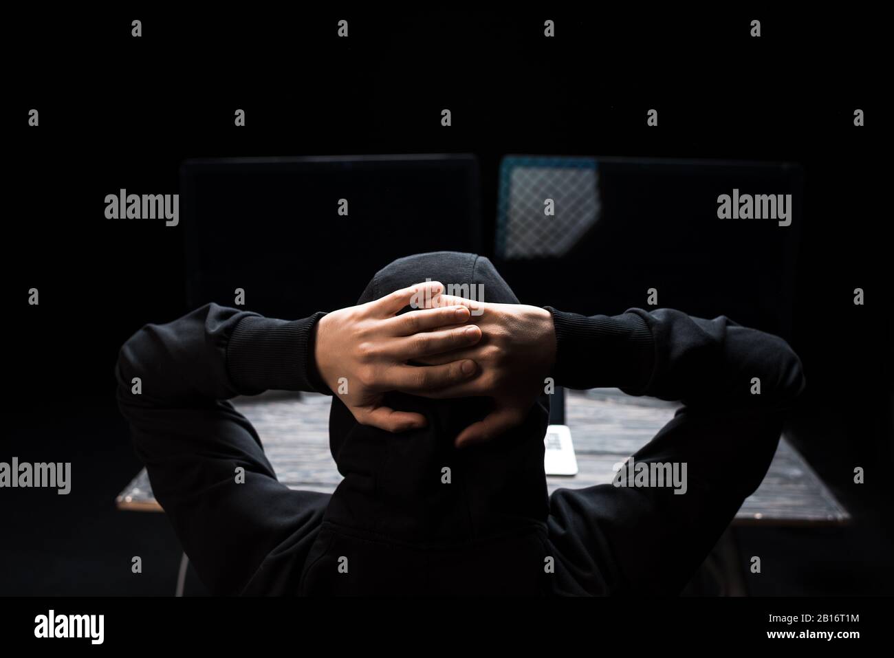 back view of hooded hacker with hands behind head near computer ...