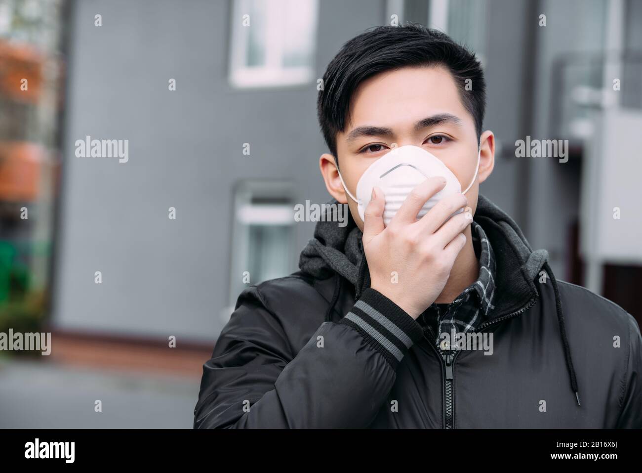 young asian man touching respirator mask and looking at camera while ...