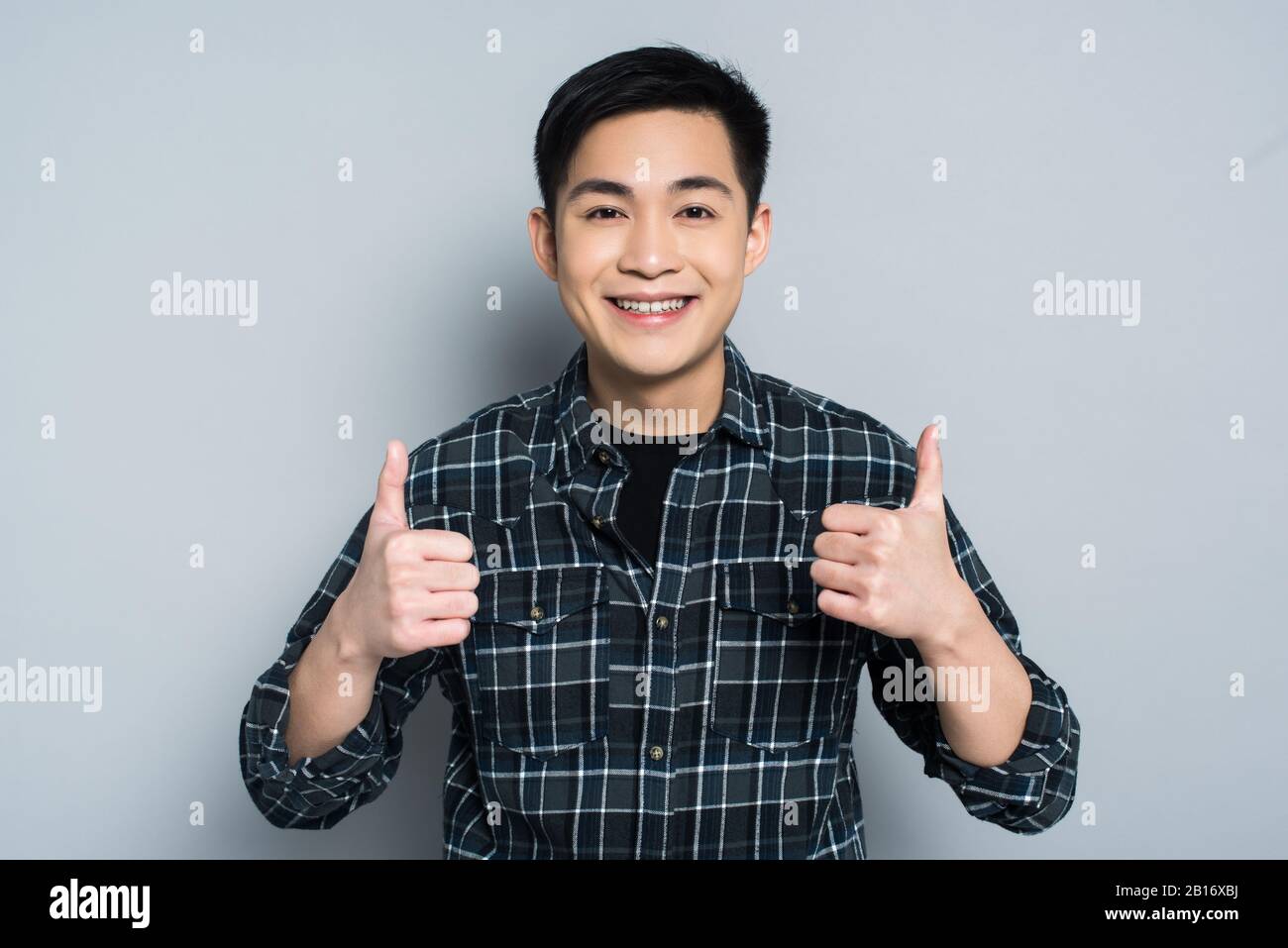 smiling asian man looking at camera while showing thumbs up on grey ...
