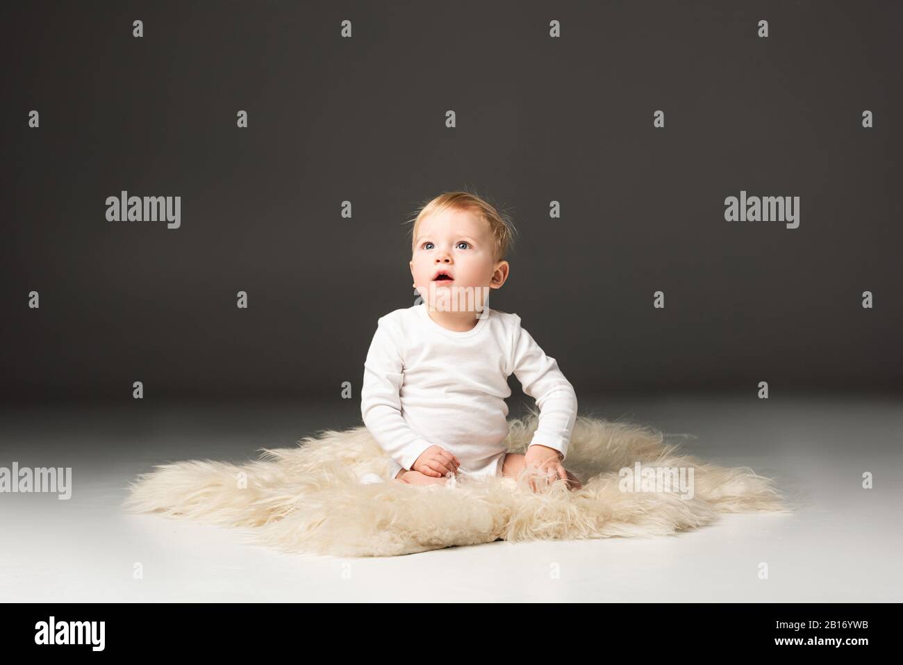 Cute kid looking up with open mouth, sitting on fur on black background ...