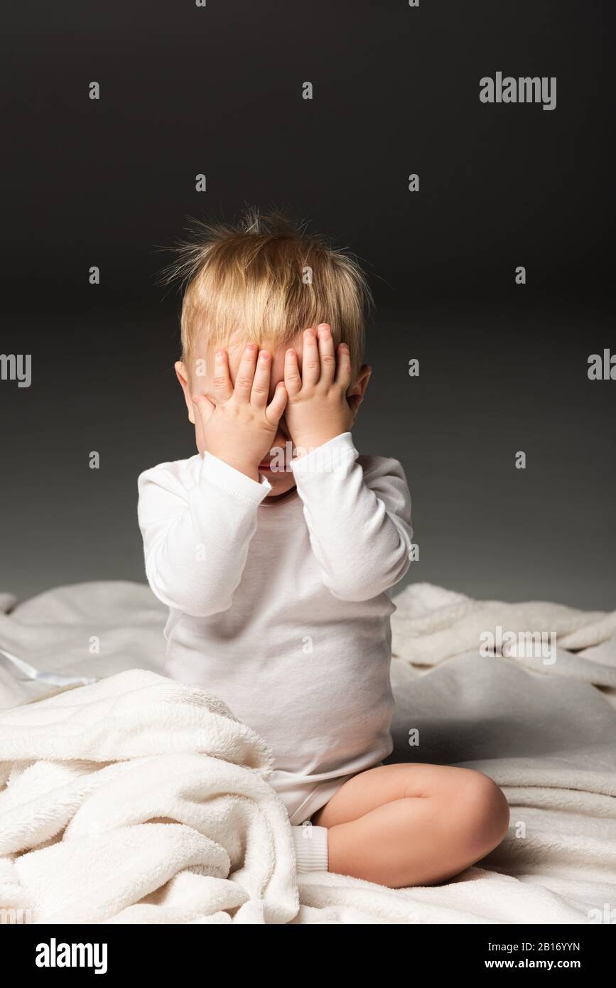 Cute child covering face with hands and sitting on blanket on black ...