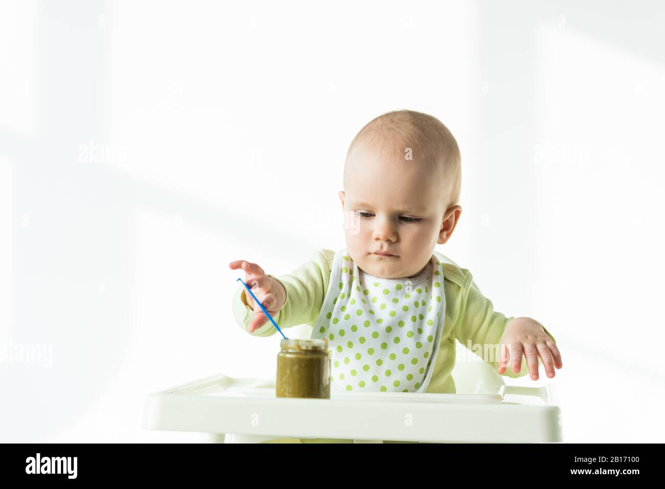 Infant holding spoon in jar of vegetable baby food on table of feeding ...