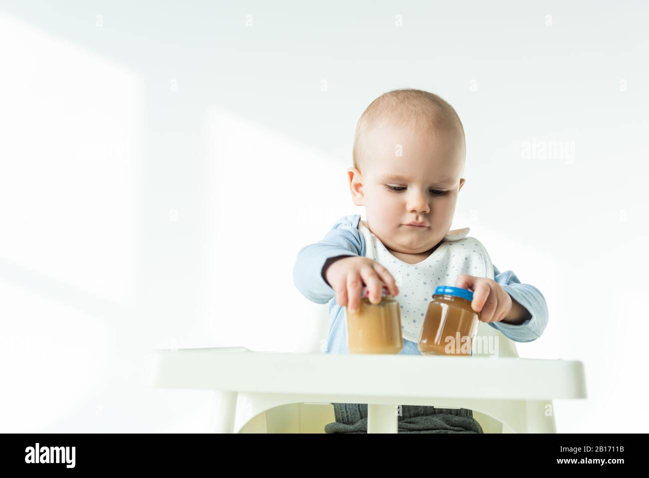 Cute baby holding jars of baby food on table of feeding chair on white ...