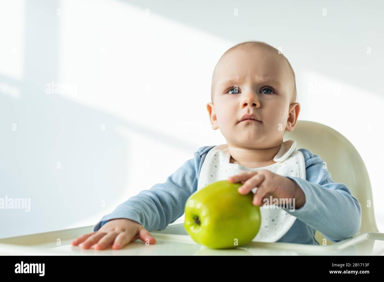 Cute baby boy touching green apple on table of feeding chair on white ...