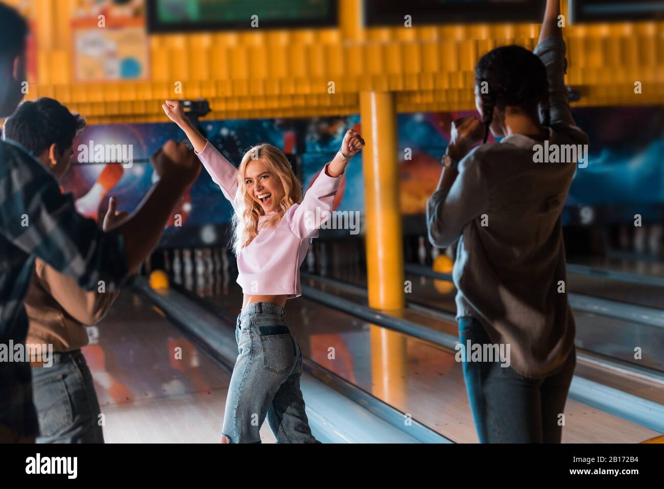 excited blonde girl showing winner gesture while standing near bowling ...