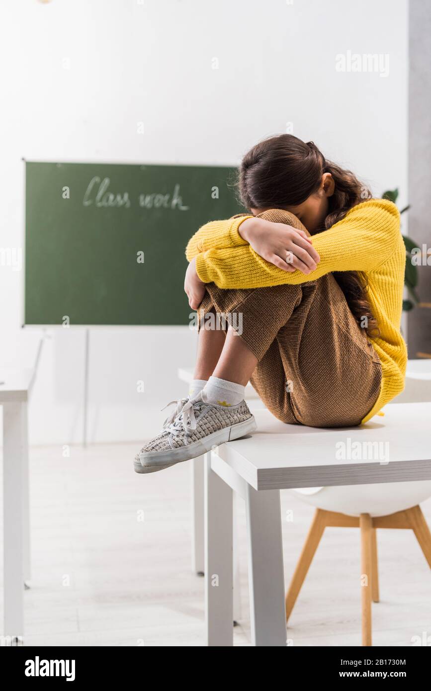 sad and bullied schoolgirl covering face while crying in classroom ...