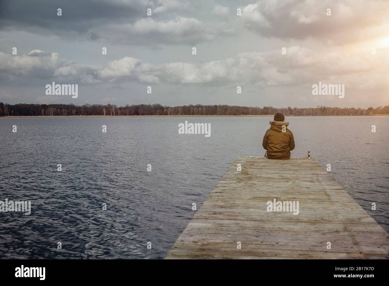 Back view of young man sitting on a jetty looking at lake in winter ...