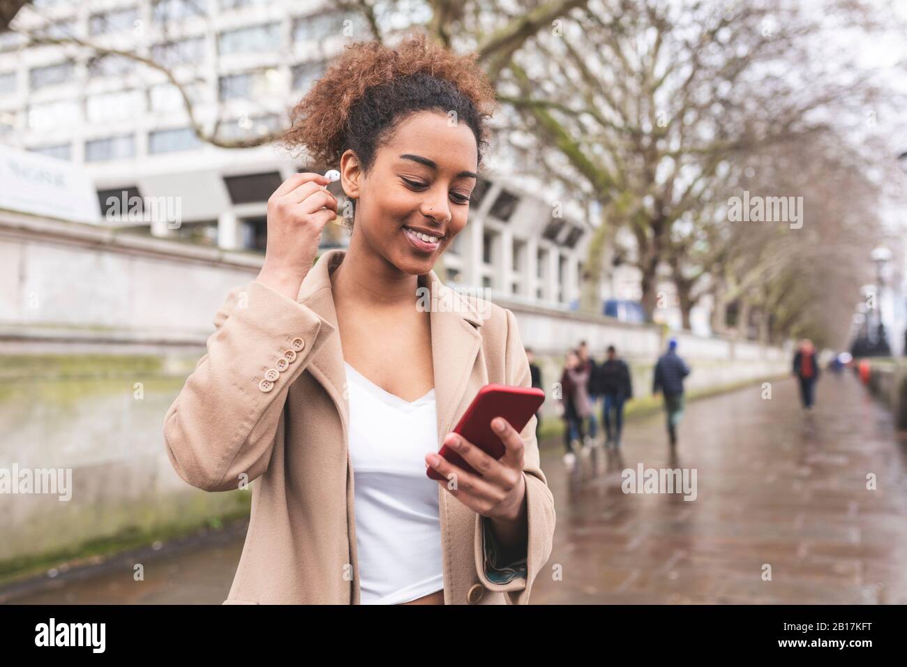 Smiling young woman with cell phone and earbuds in the city, London, UK ...
