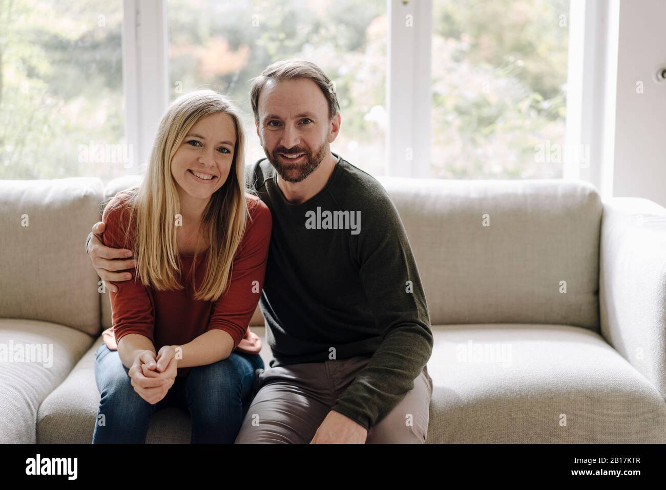 Couple sitting at home on couch, with arms around Stock Photo - Alamy