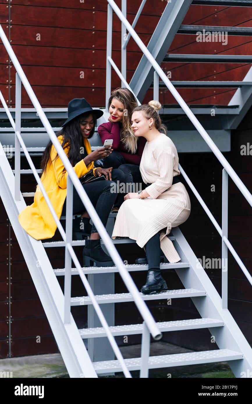 Girlfriends Sitting On Stairs Outdoors Looking At Mobile Phone Stock