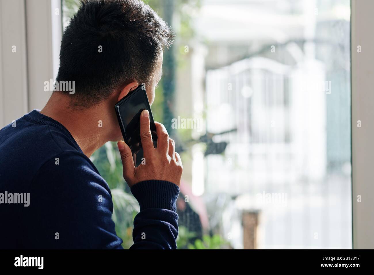 Young man looking through window when talking on phone with friend or ...