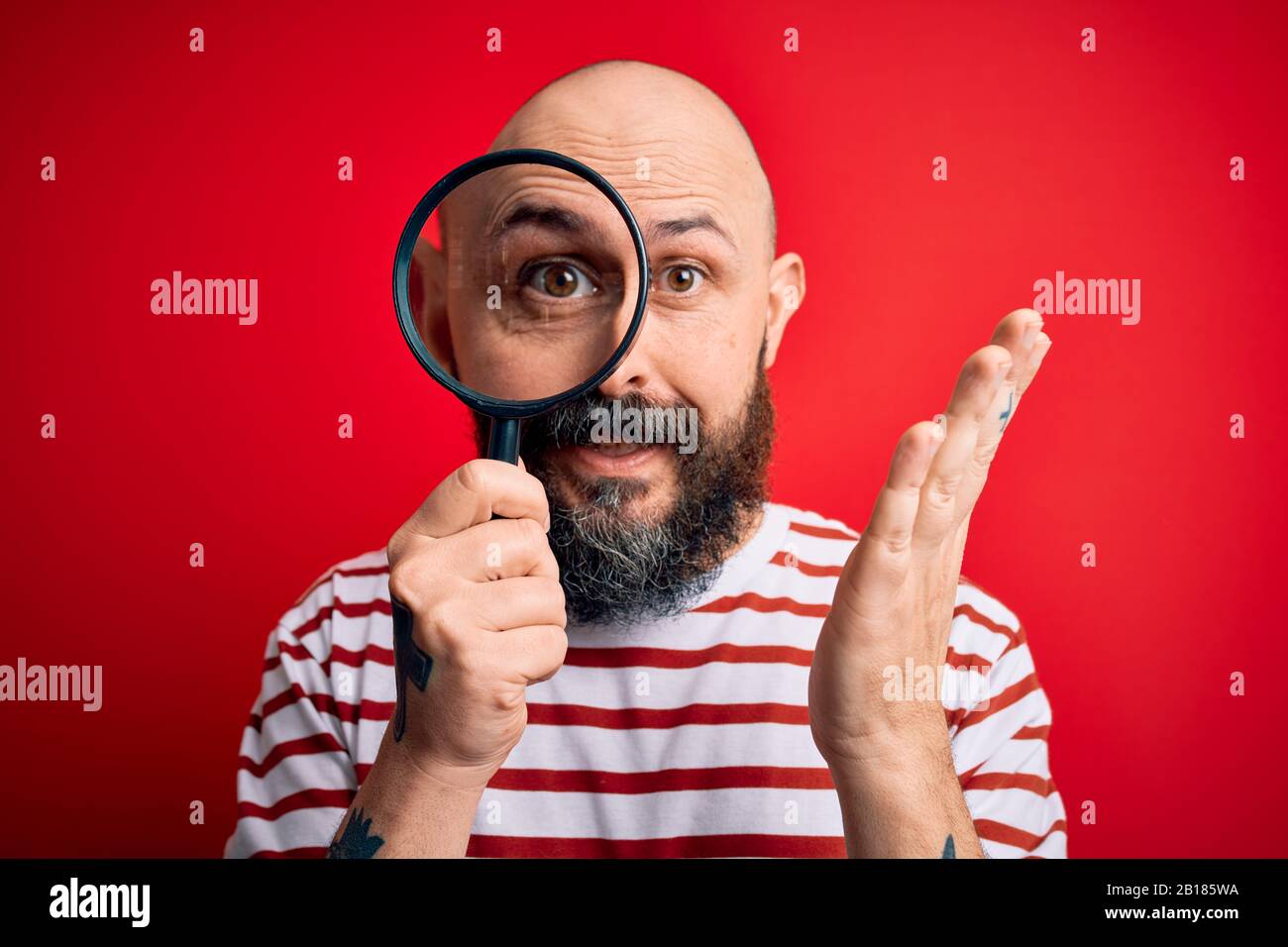Handsome detective bald man with beard using magnifying glass over red ...
