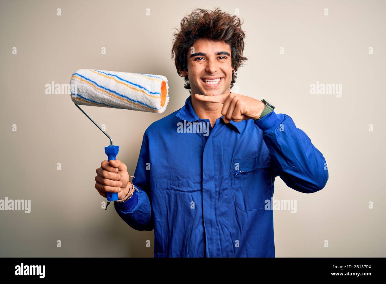 Young handsome painter man holding roller wearing uniform over isolated ...