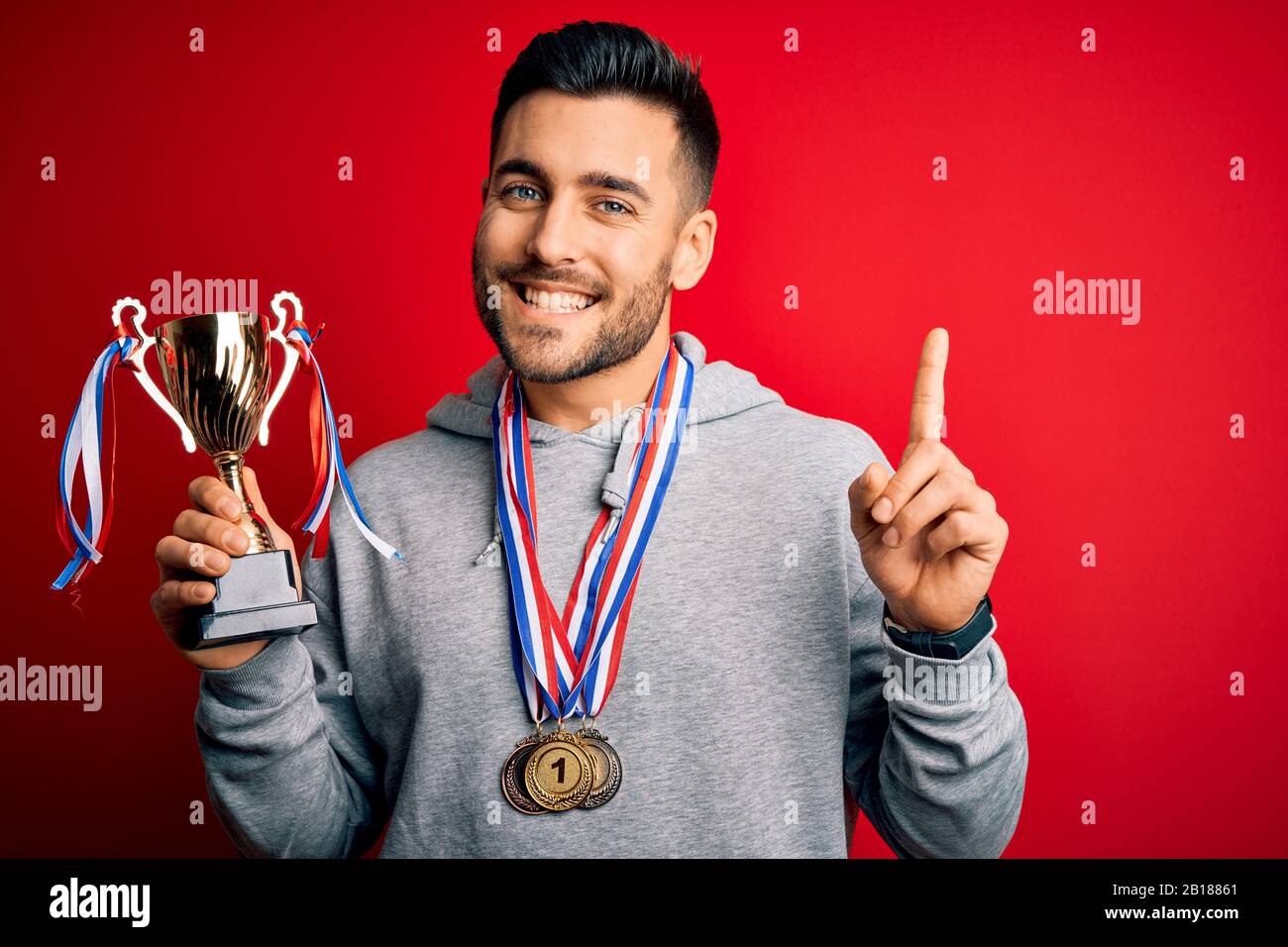 Young handsome succesful man holding trophy wearing medals over red ...