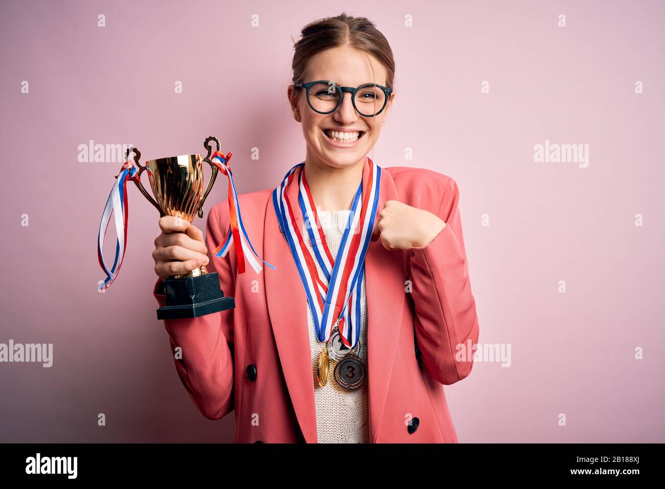 Young beautiful redhead woman holding trophy wearing medals over pink ...