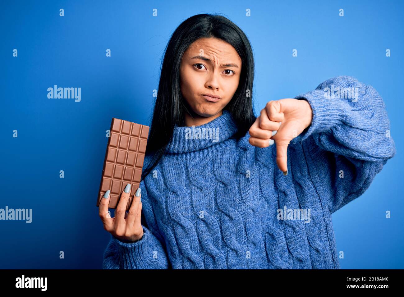 Young beautiful chinese woman holding chocolate bar over isolated blue ...