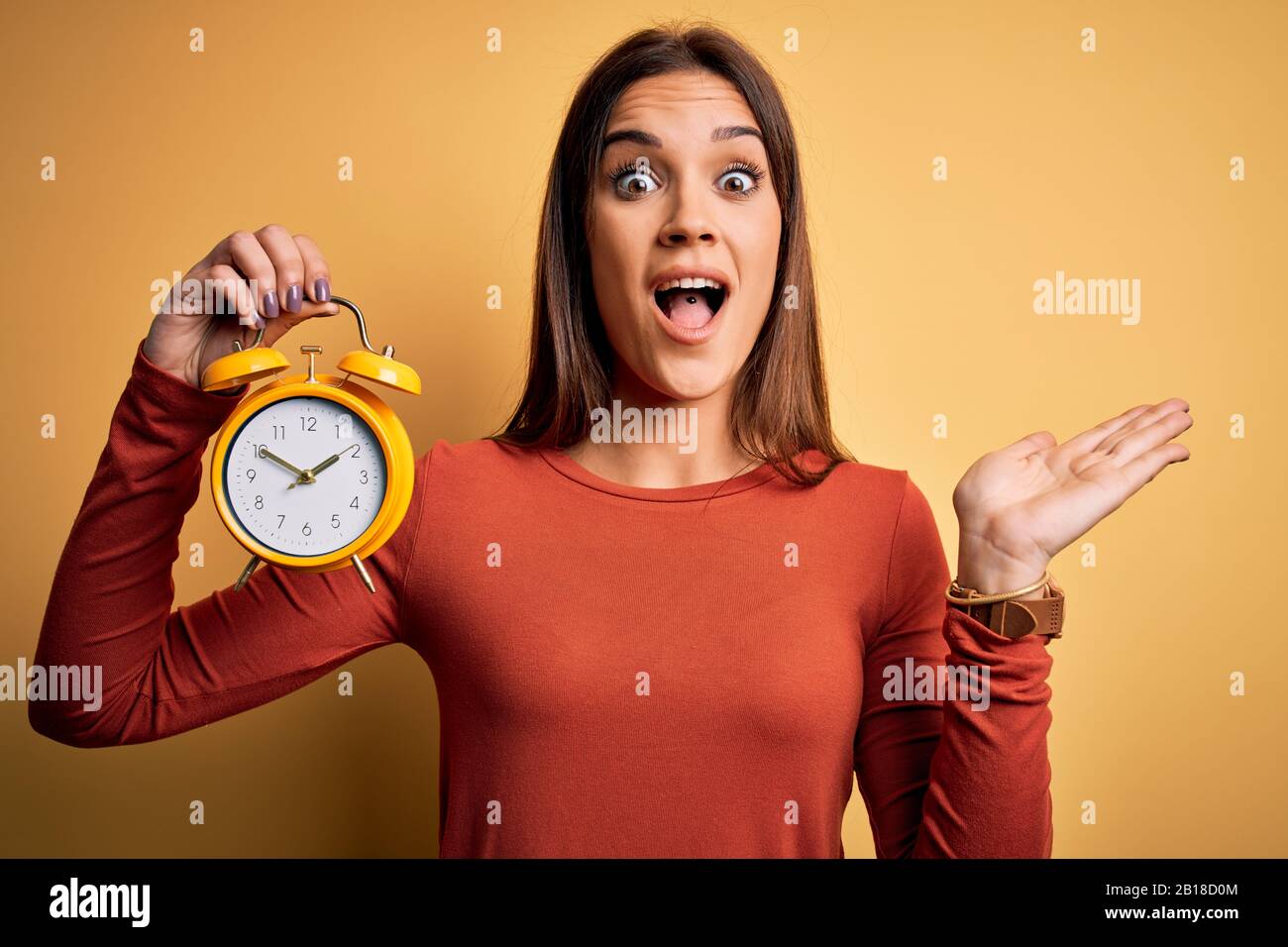 Young beautiful brunette woman holding alarm clock over isolated yellow ...